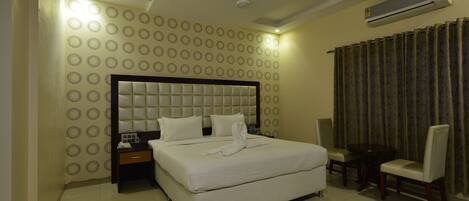 Quarto de hotel com cama grande, mesa de cabeceira, cadeira, pequena mesa e ar condicionado.