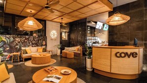 Um lobby moderno com uma receção com a placa "Cove", apresentando teto de madeira, mesas redondas de madeira e um mural com aves tropicais.