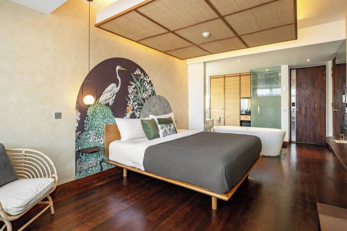 Um quarto moderno com uma cama grande, uma obra de arte na parede com um pássaro e flores, piso de madeira e área de chuveiro de vidro.
