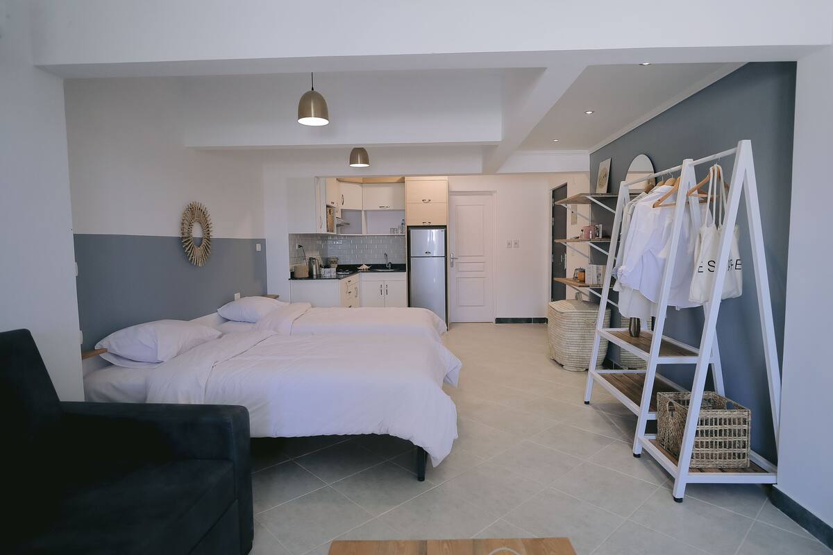 Quarto moderno e bem iluminado com uma cama, uma poltrona preta, um guarda-roupa e uma área de cozinha ao fundo.