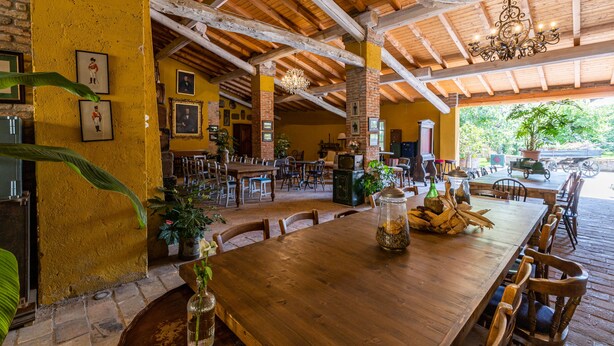 Uma ampla sala de jantar com mesas e cadeiras de madeira, um lustre e quadros nas paredes.