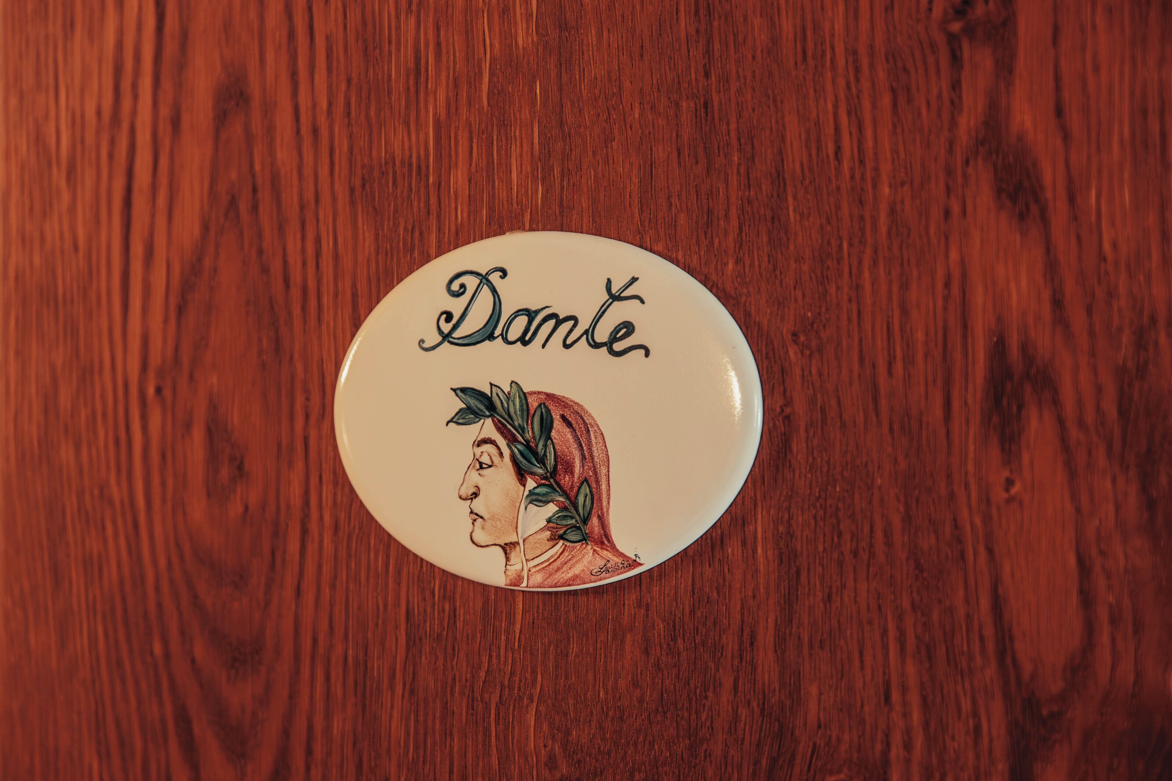 Superfície de madeira com um objeto oval branco exibindo o nome "Dante" e uma ilustração de uma pessoa com uma coroa de louros.