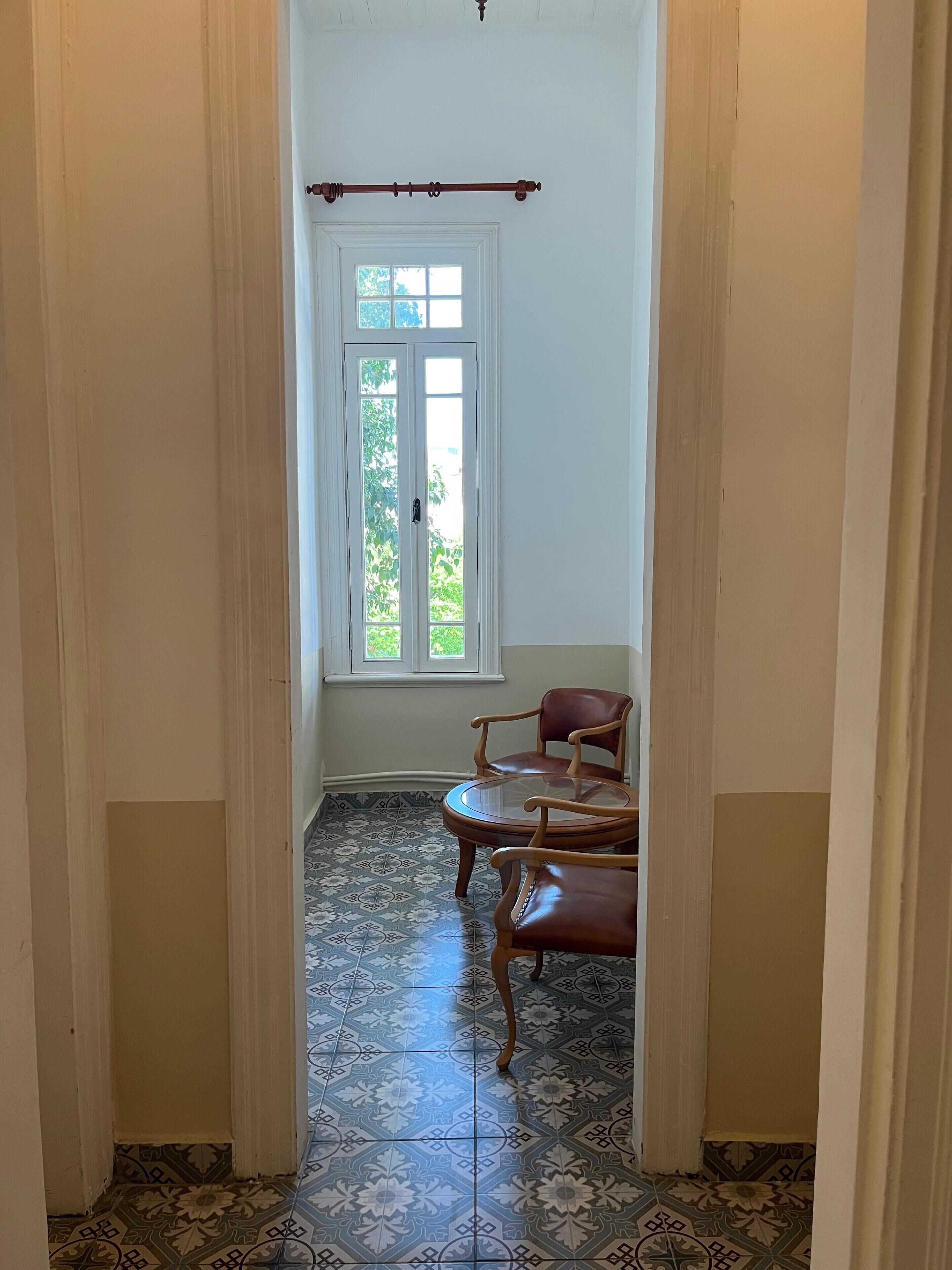 Um corredor estreito com ladrilhos estampados, uma janela com vista para vegetação e duas cadeiras de madeira.