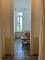 Um corredor estreito com ladrilhos estampados, uma janela com vista para vegetação e duas cadeiras de madeira.