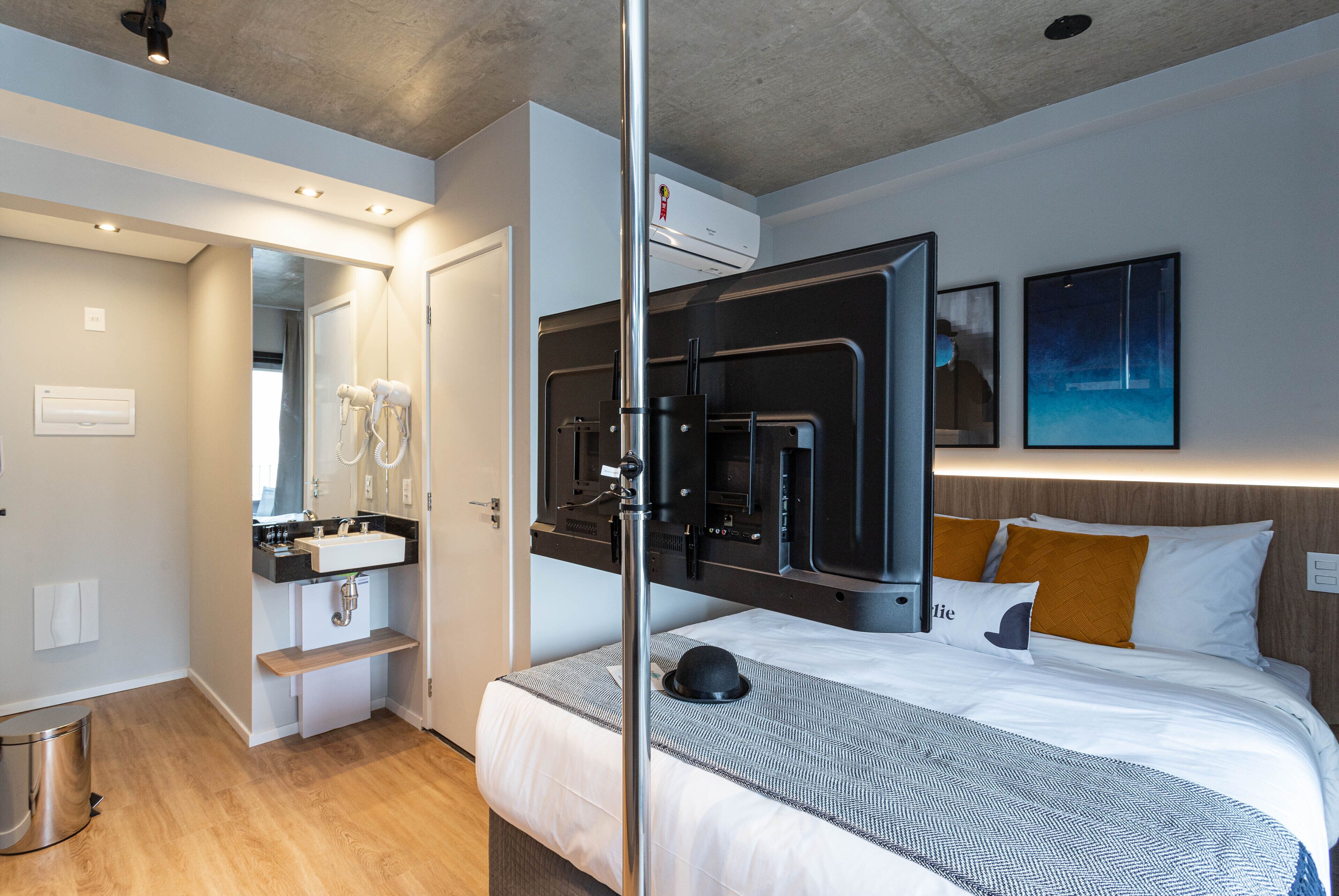 Quarto de hotel moderno com uma cama, banheiro com pia e espelho, e uma janela grande.