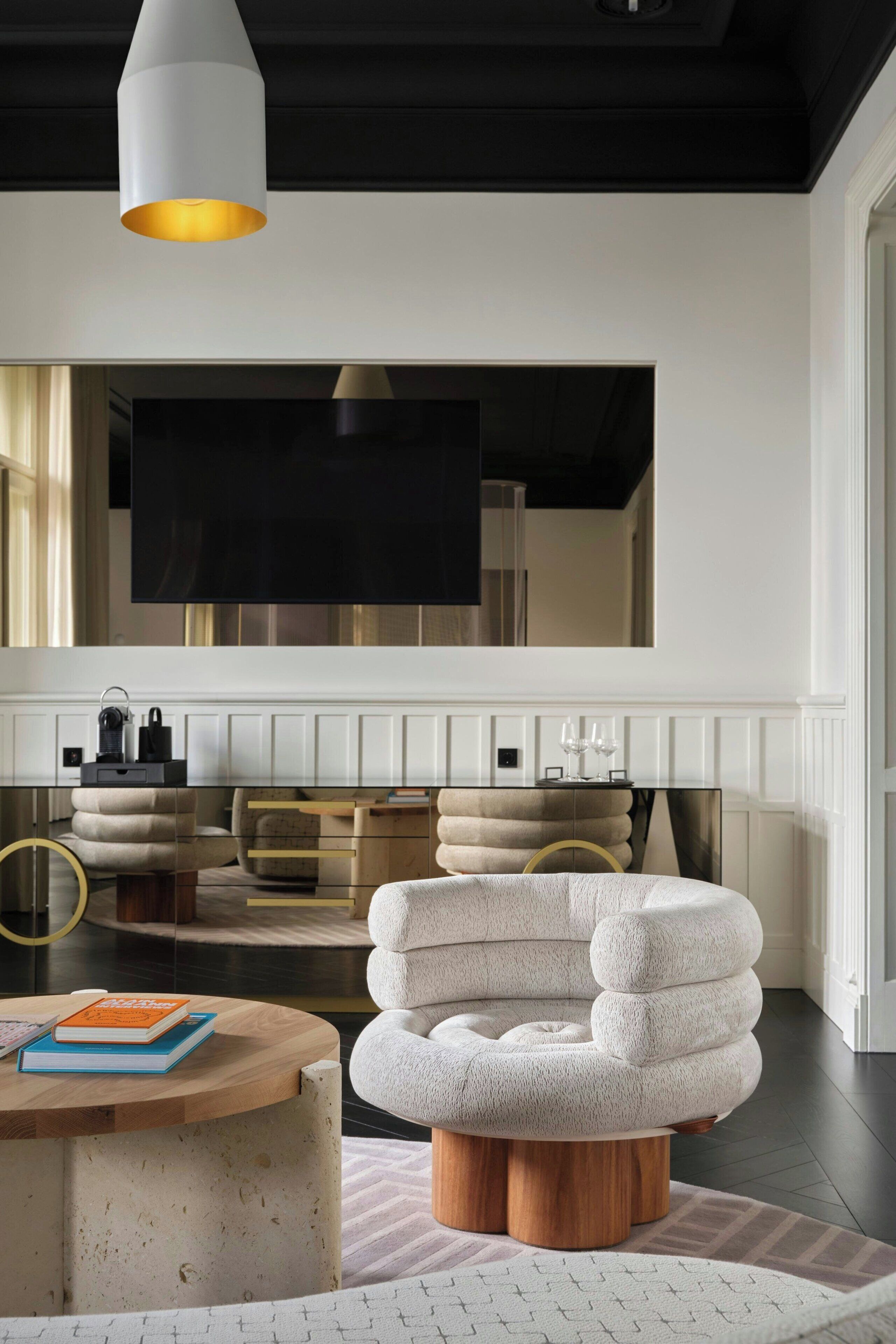 Uma sala de estar moderna com uma televisão de tela plana grande, uma mesa de centro redonda de madeira, uma cadeira em camadas diferenciada e uma luminária pendente.