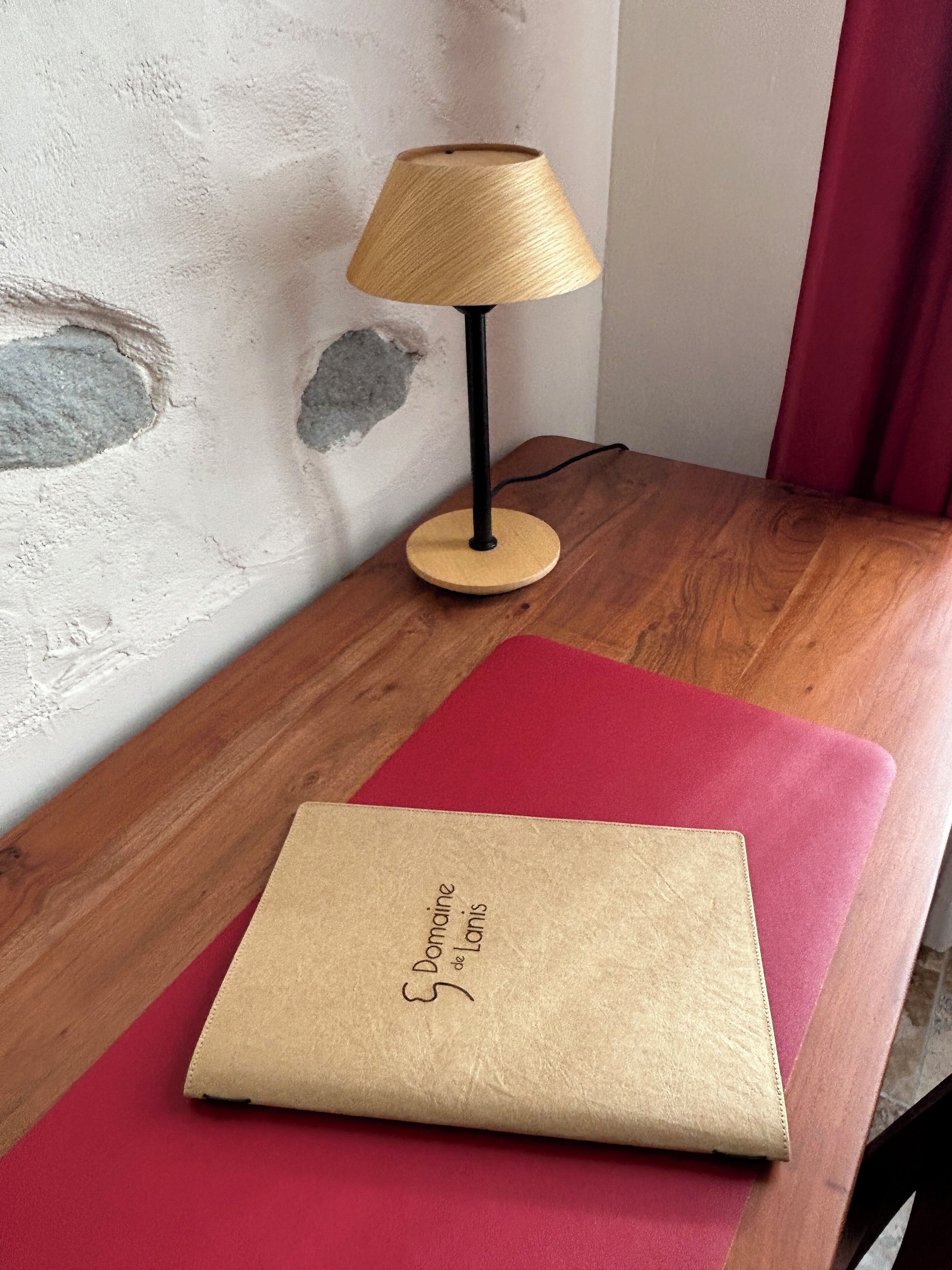 Uma mesa de madeira com um tapete vermelho, uma lâmpada e um caderno bege com texto manuscrito.