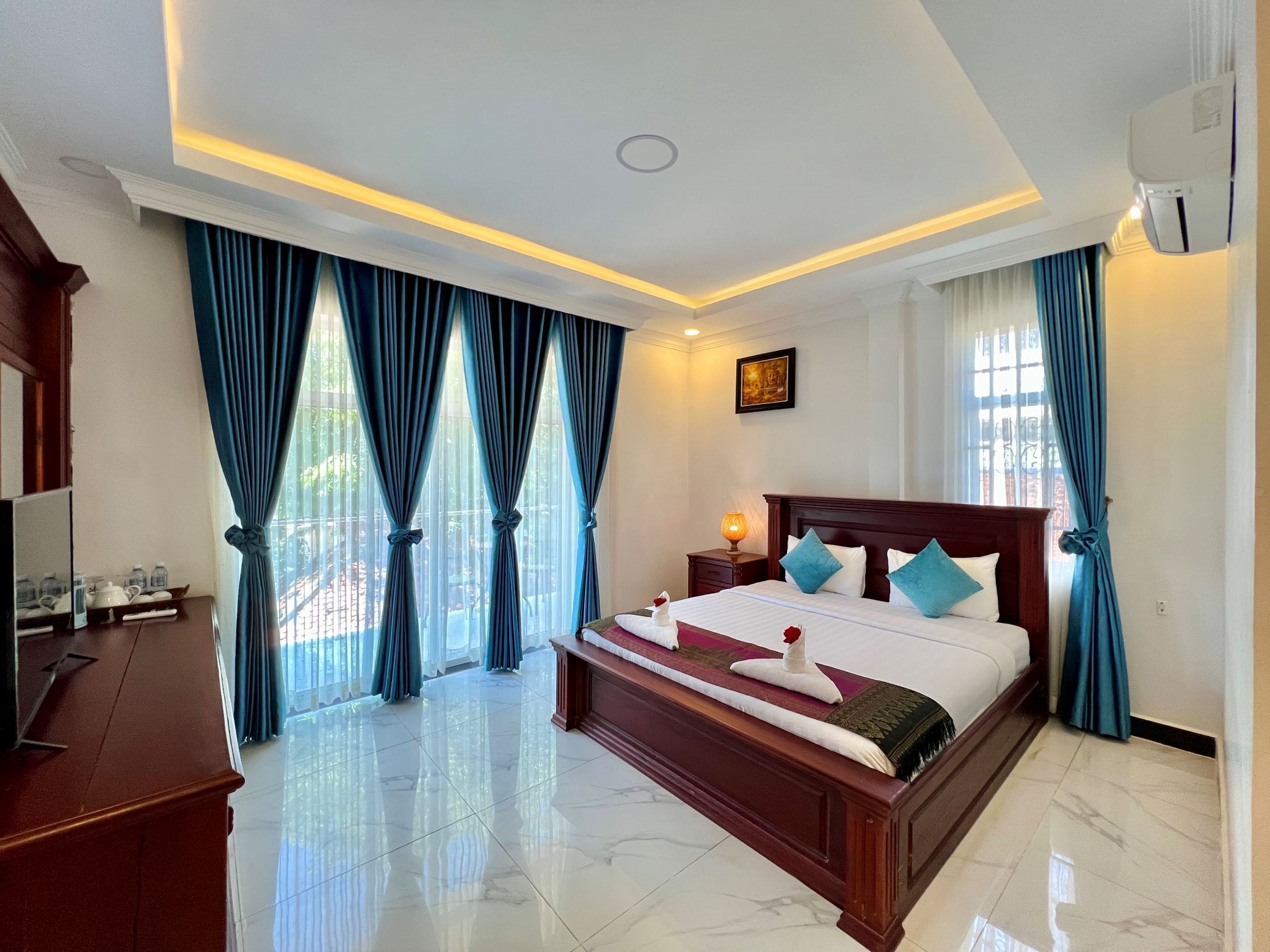 Quarto de hotel com uma cama grande, cômoda de madeira, escrivaninha com luminária e uma janela ampla com cortinas.