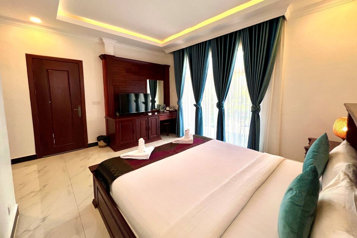 Quarto de hotel com uma cama grande, cômoda de madeira, televisão e janela com cortinas.