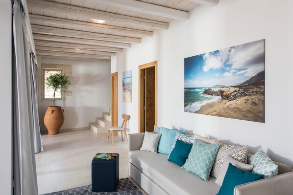 Uma sala de estar moderna com um sofá bege, almofadas estampadas, uma grande pintura de praia e uma planta em vaso.