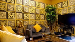 Uma sala de estar com sofá, mesa de centro e televisão. As paredes apresentam padrões elaborados.