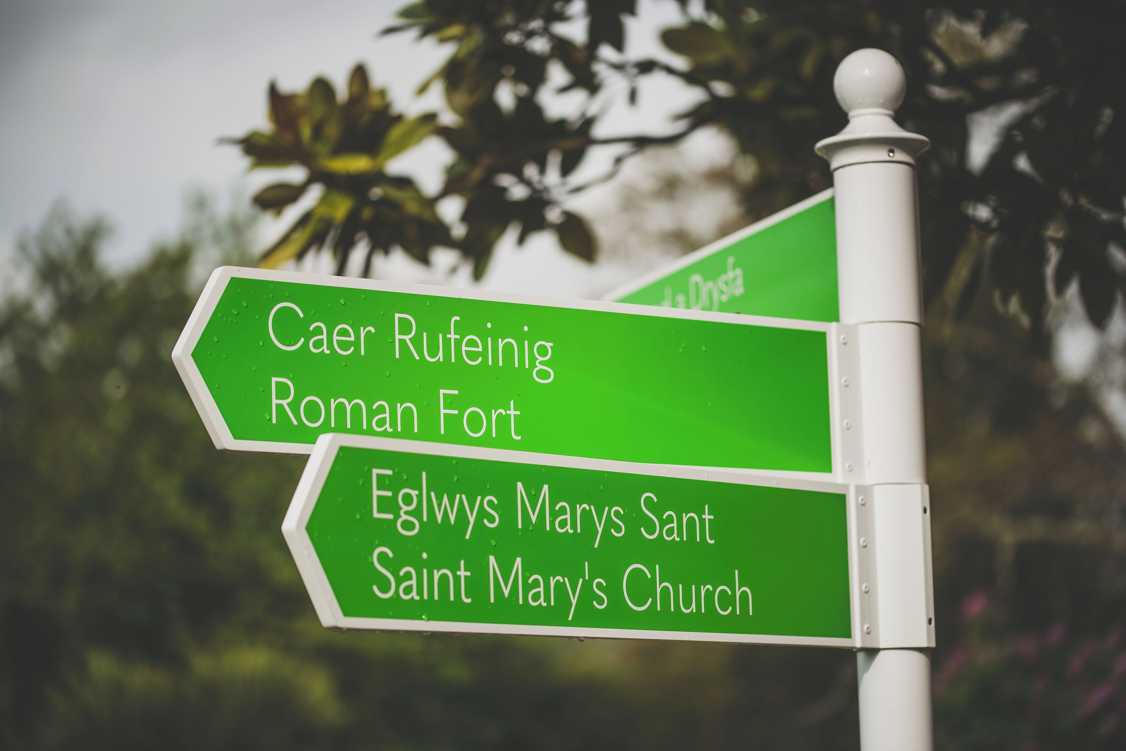 Placa de sinalização com painéis verdes indicando direções para o Forte Romano de Caer Rufeinig e a Igreja de Eglwys Marys Sant Saint Mary's.