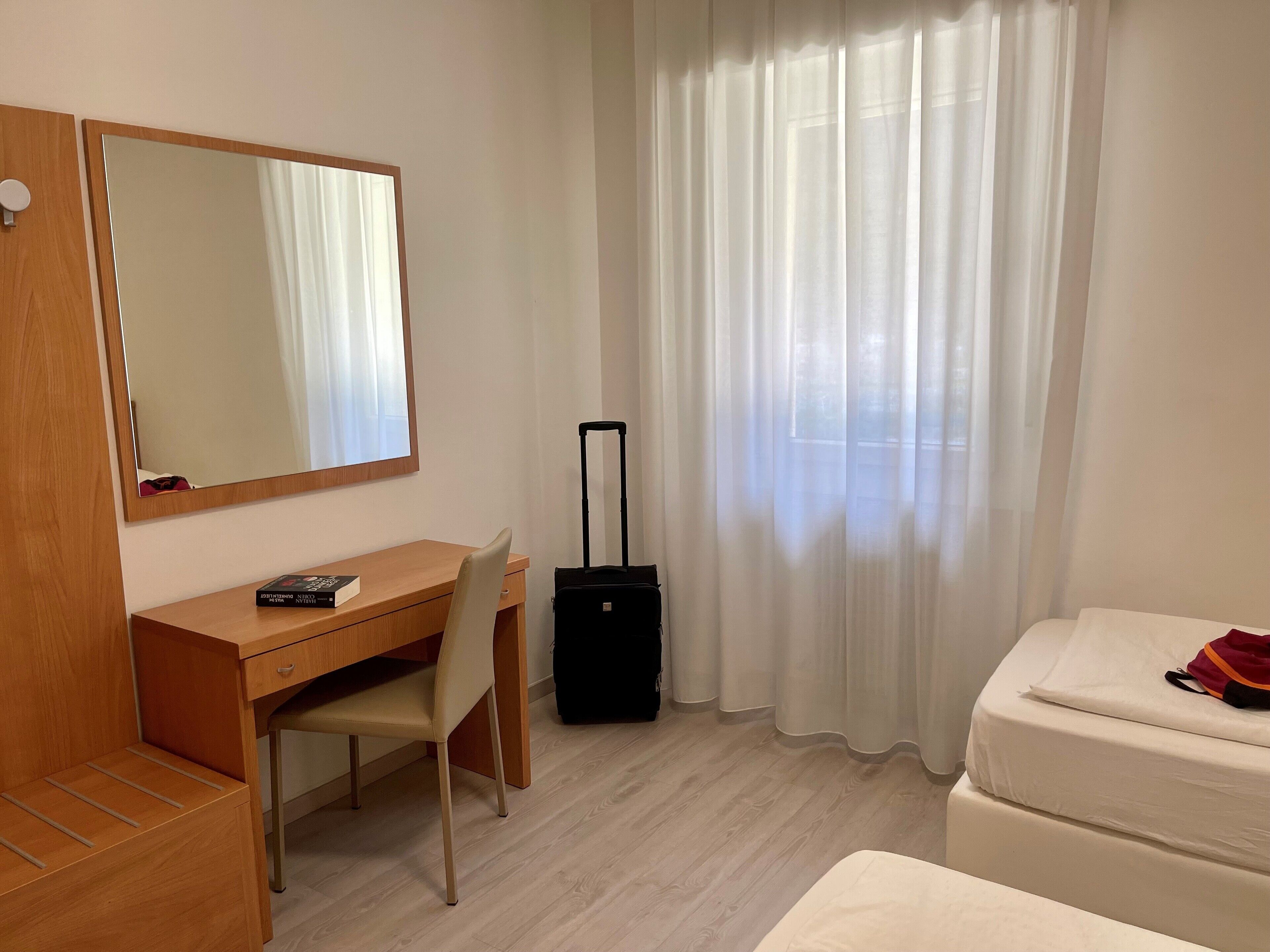 Quarto de hotel com uma escrivaninha de madeira, uma cadeira, uma cama, uma mala e uma janela com cortinas leves.