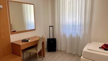 Quarto de hotel com uma escrivaninha de madeira, uma cadeira, uma cama, uma mala e uma janela com cortinas leves.