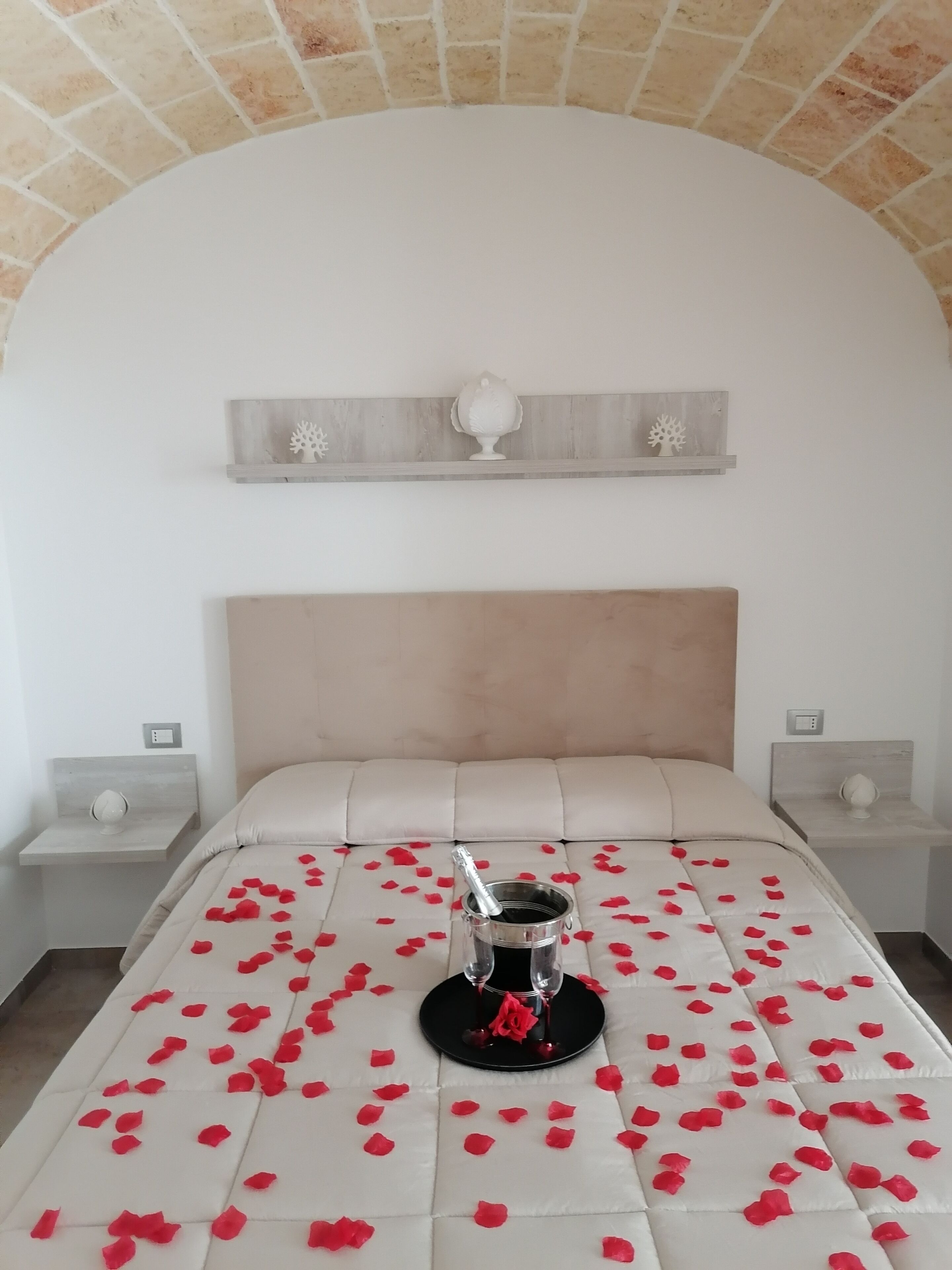 Quarto de hotel com uma cama coberta de pétalas de rosa, uma mesa de cabeceira com uma garrafa e uma estante decorativa acima da cama.
