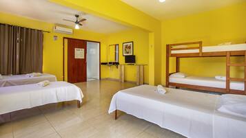 Quarto de hotel com duas camas de solteiro, uma cama beliche, uma escrivaninha com cadeira, um ventilador de teto, um ar condicionado e uma porta com placa.