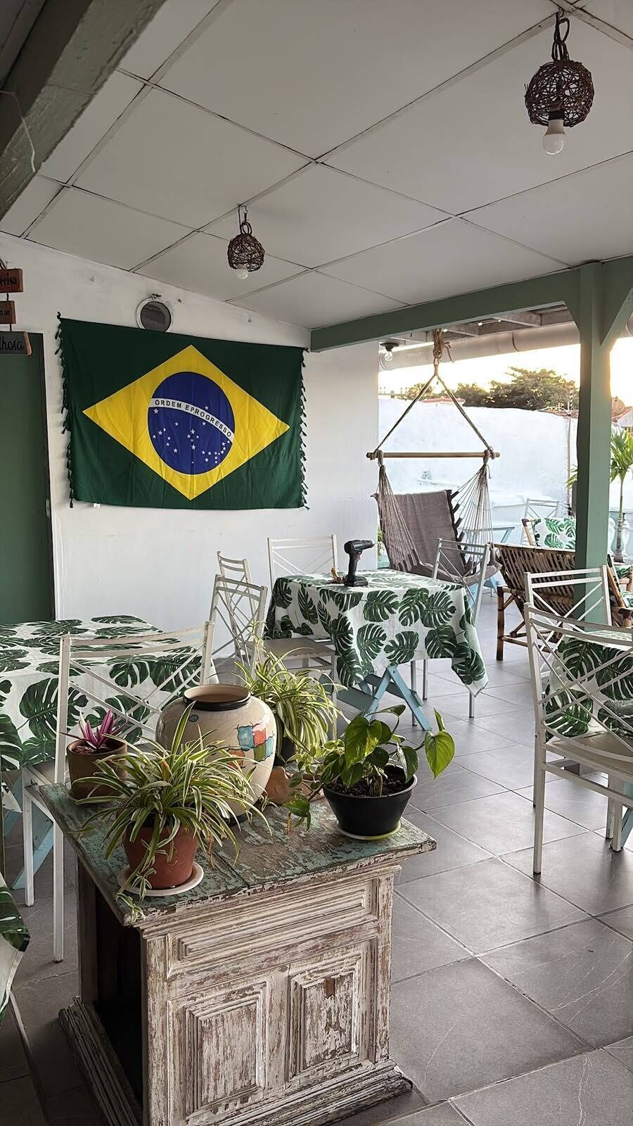 Um quarto com uma bandeira do Brasil, uma rede, uma mesa com toalha de mesa de folha tropical e vários vasos de plantas.