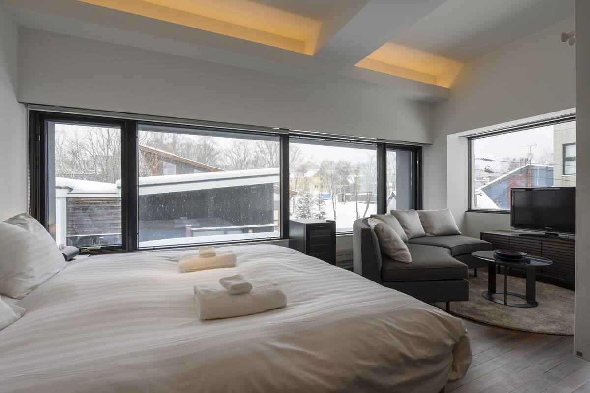 Quarto de hotel moderno com uma cama grande, um sofá, uma TV e vista para o exterior.