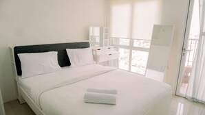 Uma cama bem arrumada com lençóis brancos e uma toalha dobrada, uma mesa de cabeceira com espelho, e vista para o exterior por uma grande janela.