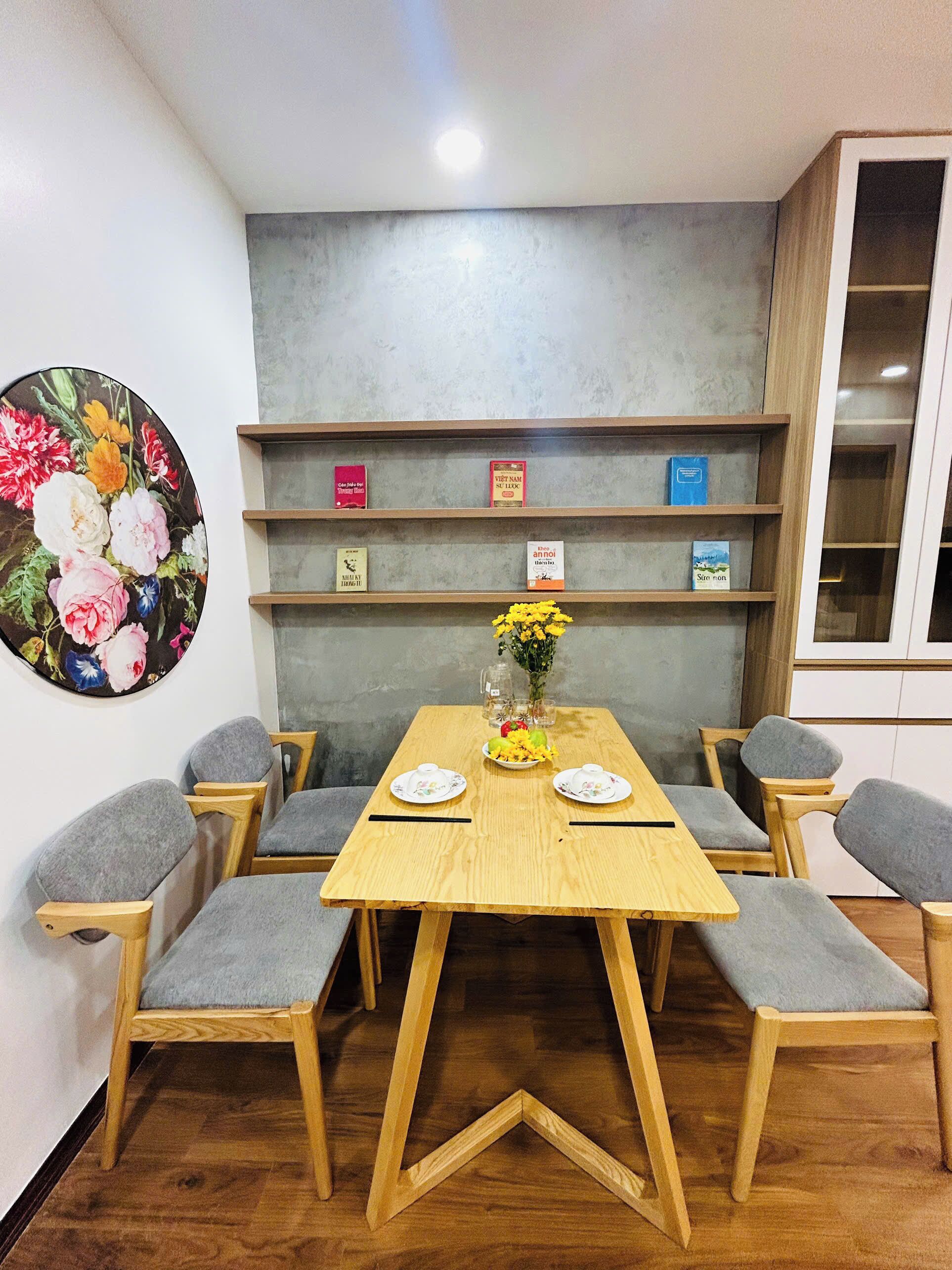 Uma área de refeições com uma mesa de madeira e cadeiras estofadas em cinza, rodeada por estantes com livros e um quadro floral.