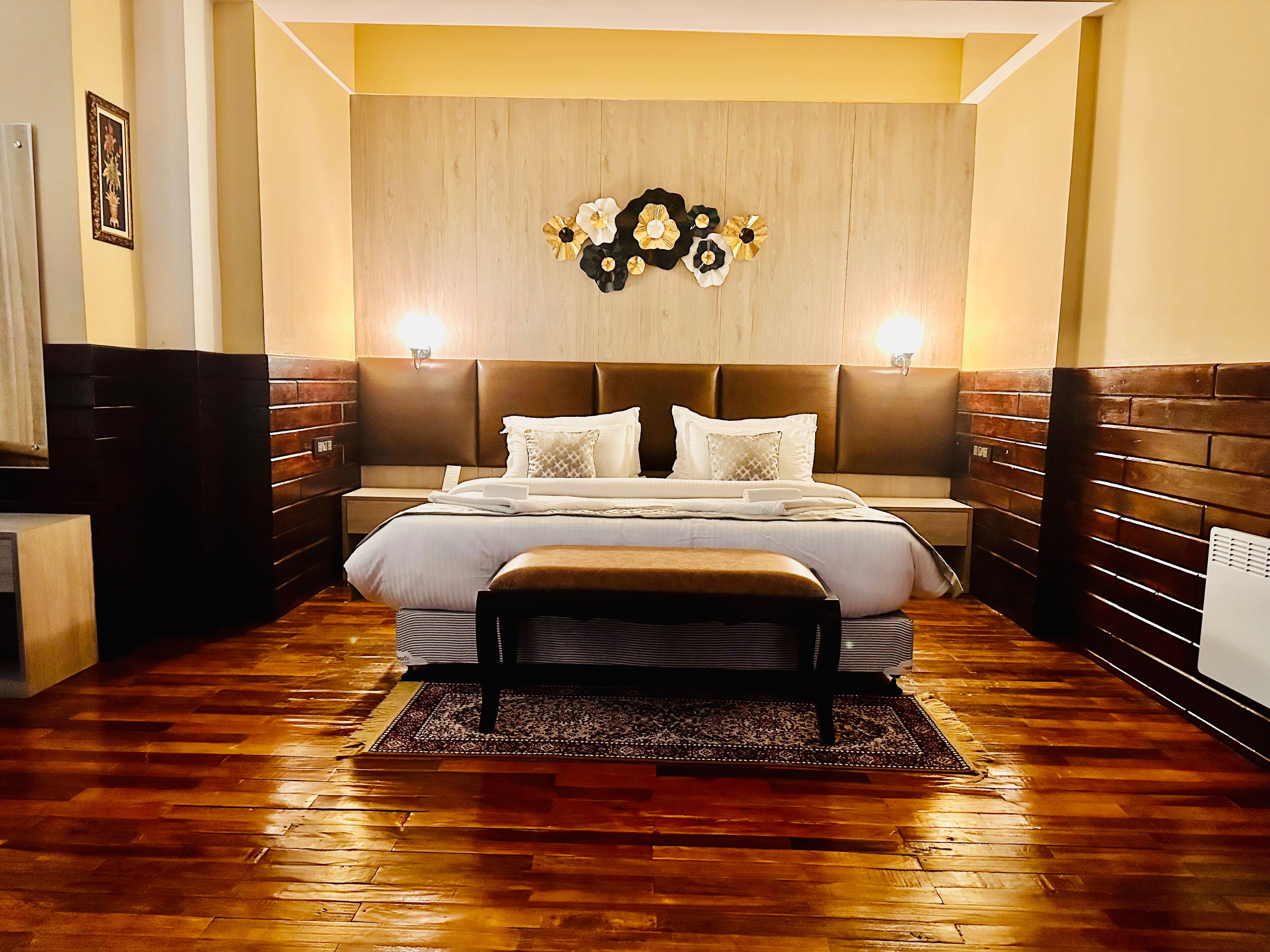 Quarto com piso de madeira, cama com roupa de cama branca, duas luminárias de cabeceira e um objeto decorativo na parede acima da cama.