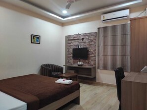 Quarto de hotel com uma cama, uma televisão presa a uma parede de pedra, uma escrivaninha, uma cadeira e um aparelho de ar condicionado.