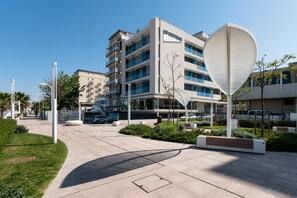 Um hotel moderno com uma placa de 'Sporting' bem visível, apresentando uma estrutura em vários andares com varandas, um pátio ajardinado e uma escultura branca singular.