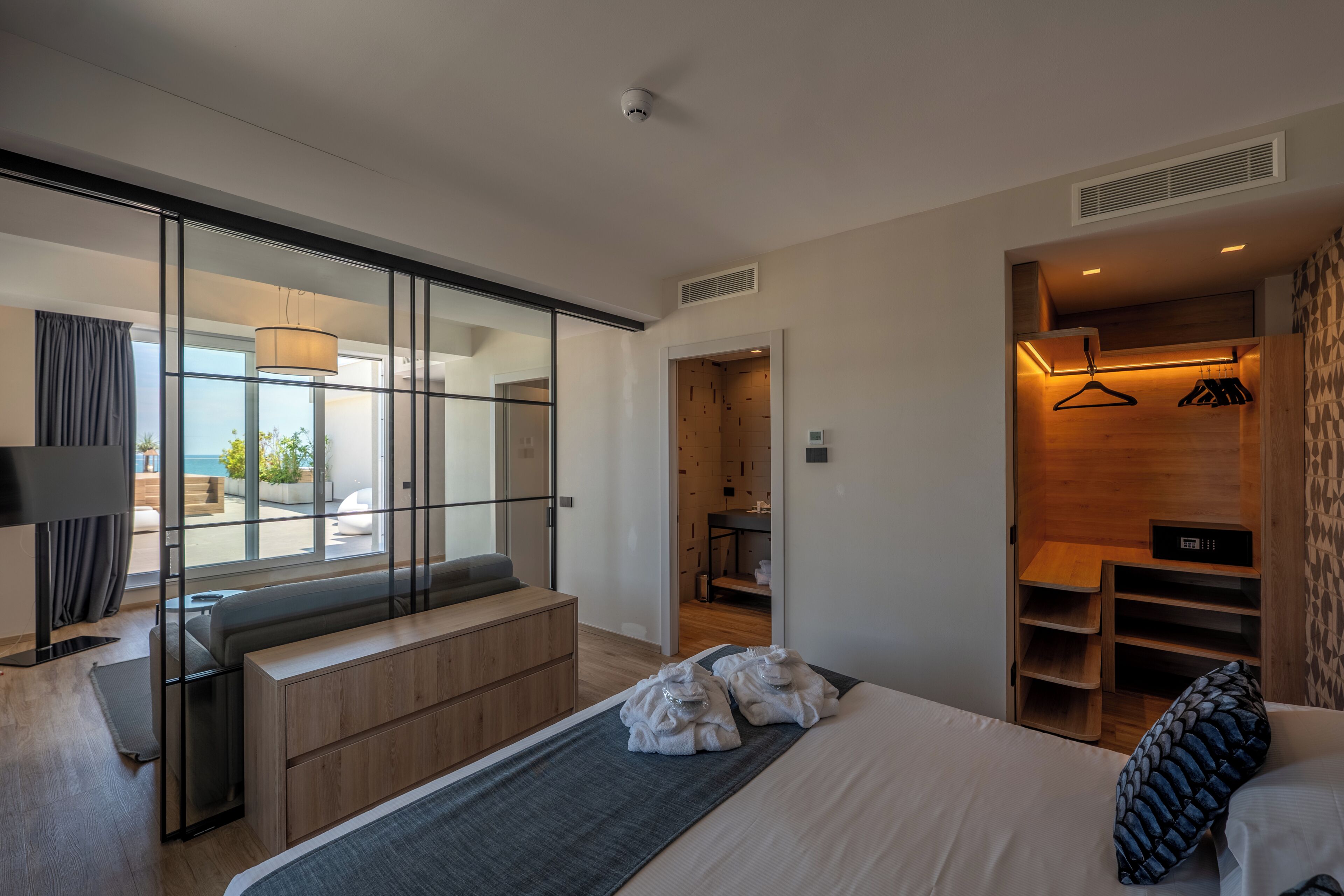 Quarto de hotel moderno com uma cama grande, área de estar, closet espaçoso e vista através de um painel de vidro.