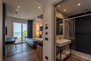 Quarto de hotel moderno com uma cama grande, uma mesa de escritório, vista para o mar e banheiro separado com chuveiro de vidro.