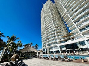 Um hotel moderno com piscina, palmeiras e uma estrutura com telhado de palha.