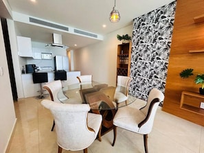 Uma sala de jantar moderna com uma mesa de vidro rodeada por cadeiras estofadas, um aparador de madeira e uma cortina estampada.