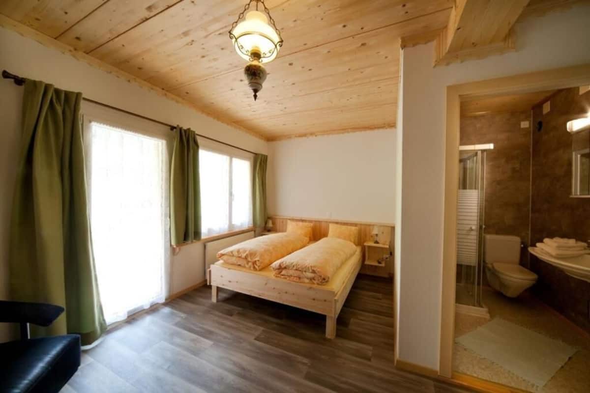 Quarto com cama de madeira, mesa de cabeceira, janela com cortinas e banheiro com vaso sanitário e pia.
