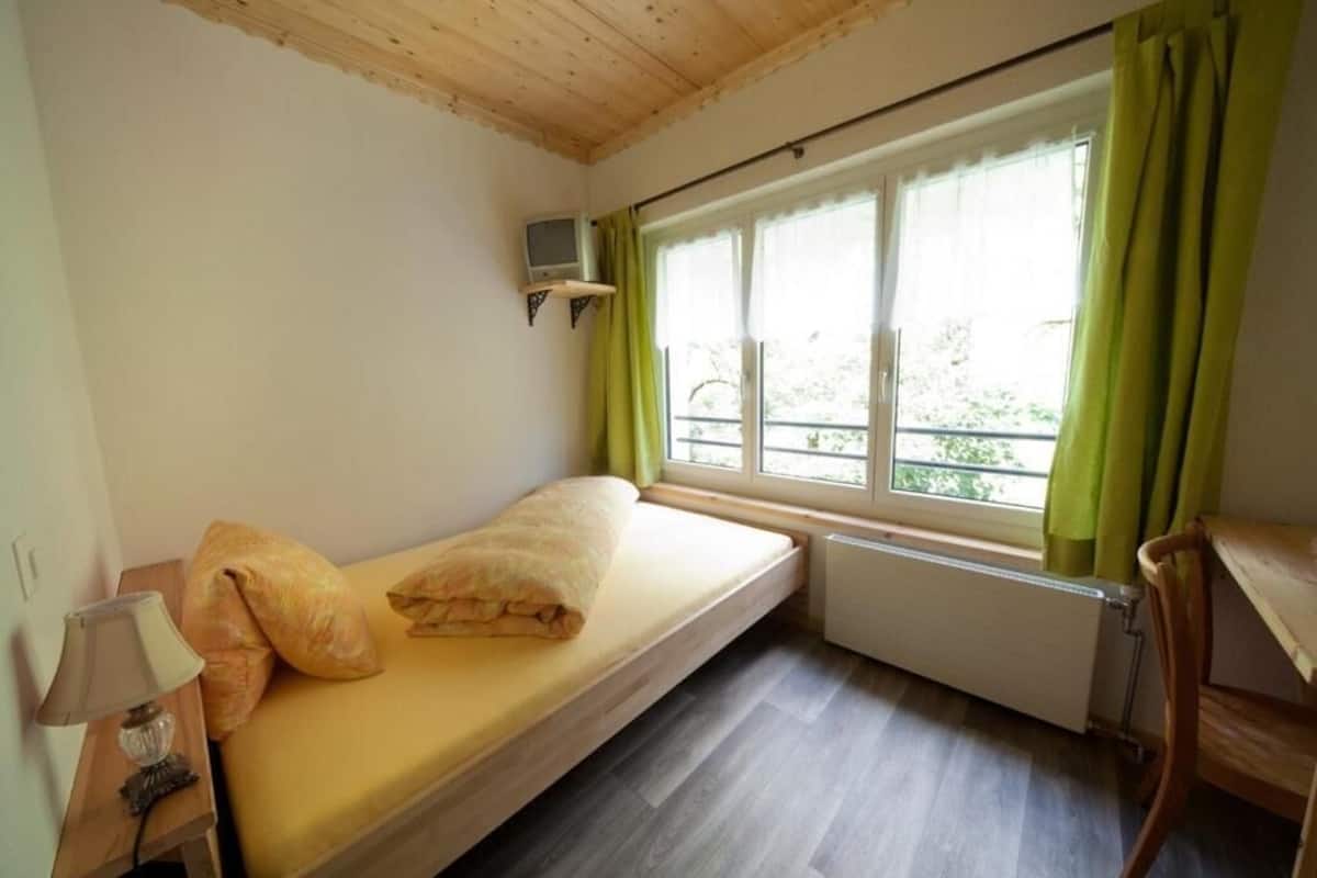 Quarto pequeno com uma cama de solteiro, uma mesa, uma cadeira, janela com cortinas e uma televisão em uma prateleira.