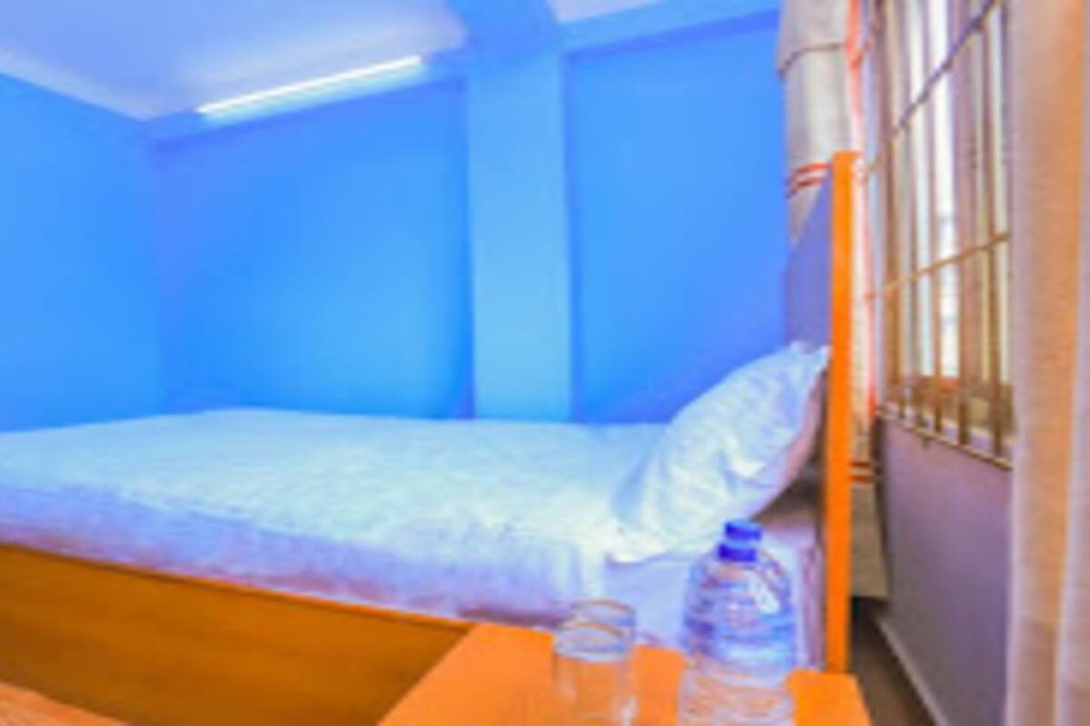 Quarto pequeno com cama de solteiro, estrutura de madeira, mesa de cabeceira com uma garrafa de água e janela com grades.