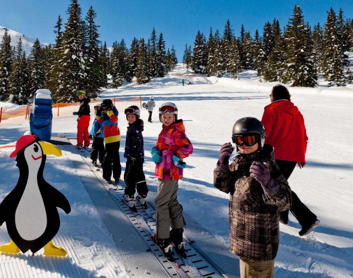 Um grupo de pessoas numa encosta nevada para esquiar, com um boneco de pinguim.