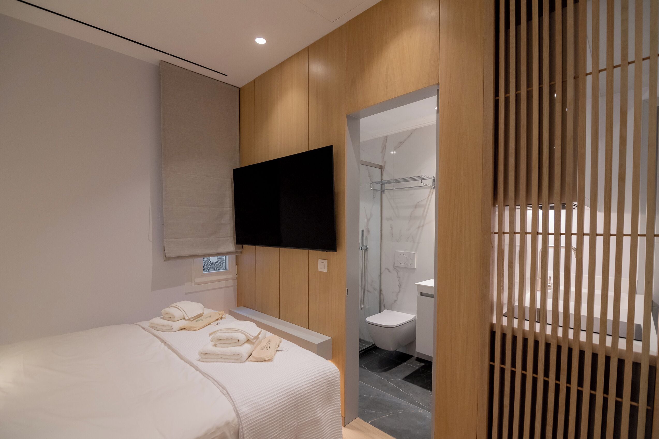 Quarto moderno com cama, televisão de tela plana, parede revestida em madeira, banheiro com box de vidro e vaso sanitário.
