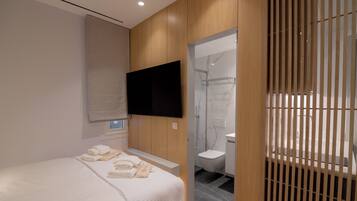Quarto moderno com cama, televisão de tela plana, parede revestida em madeira, banheiro com box de vidro e vaso sanitário.