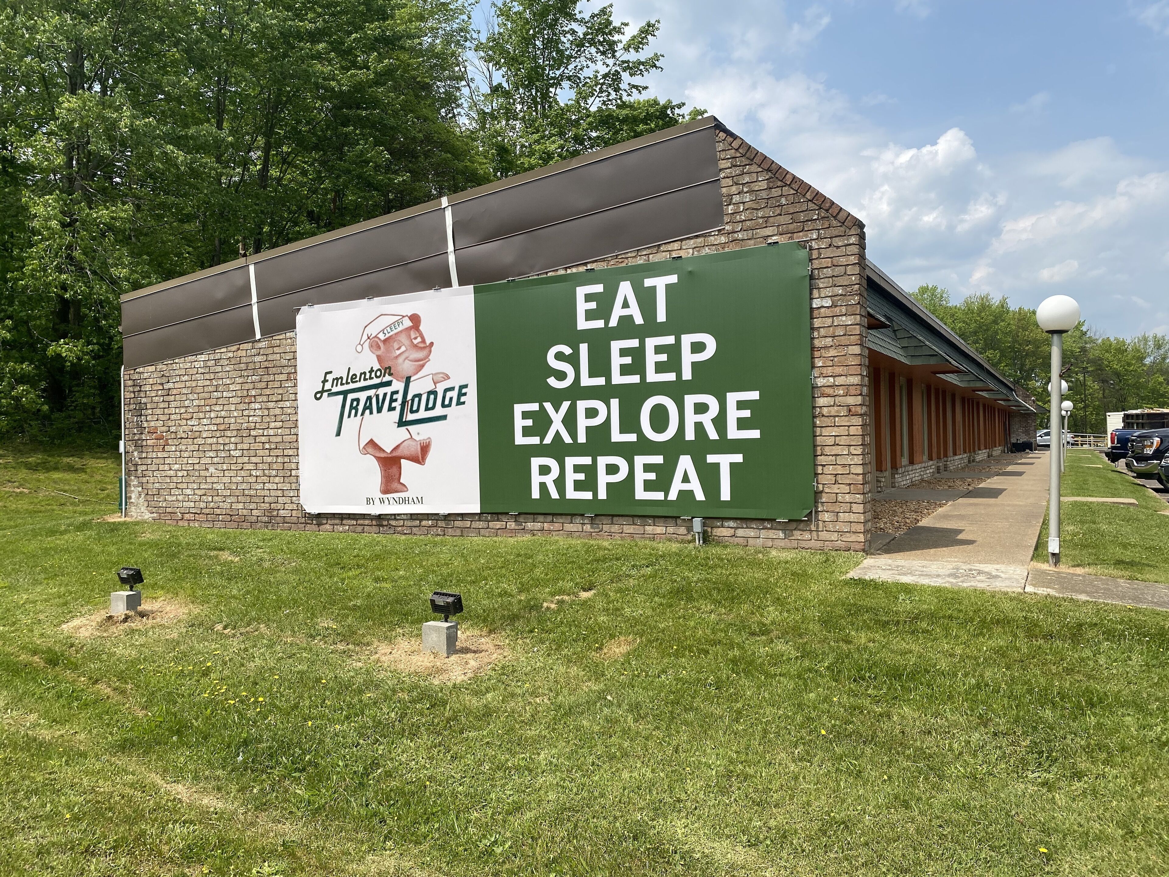 Um edifício de tijolos com uma grande placa anunciando 'EAT SLEEP EXPLORE REPEAT' e uma placa menor para 'Emleton Travel Lodge'.