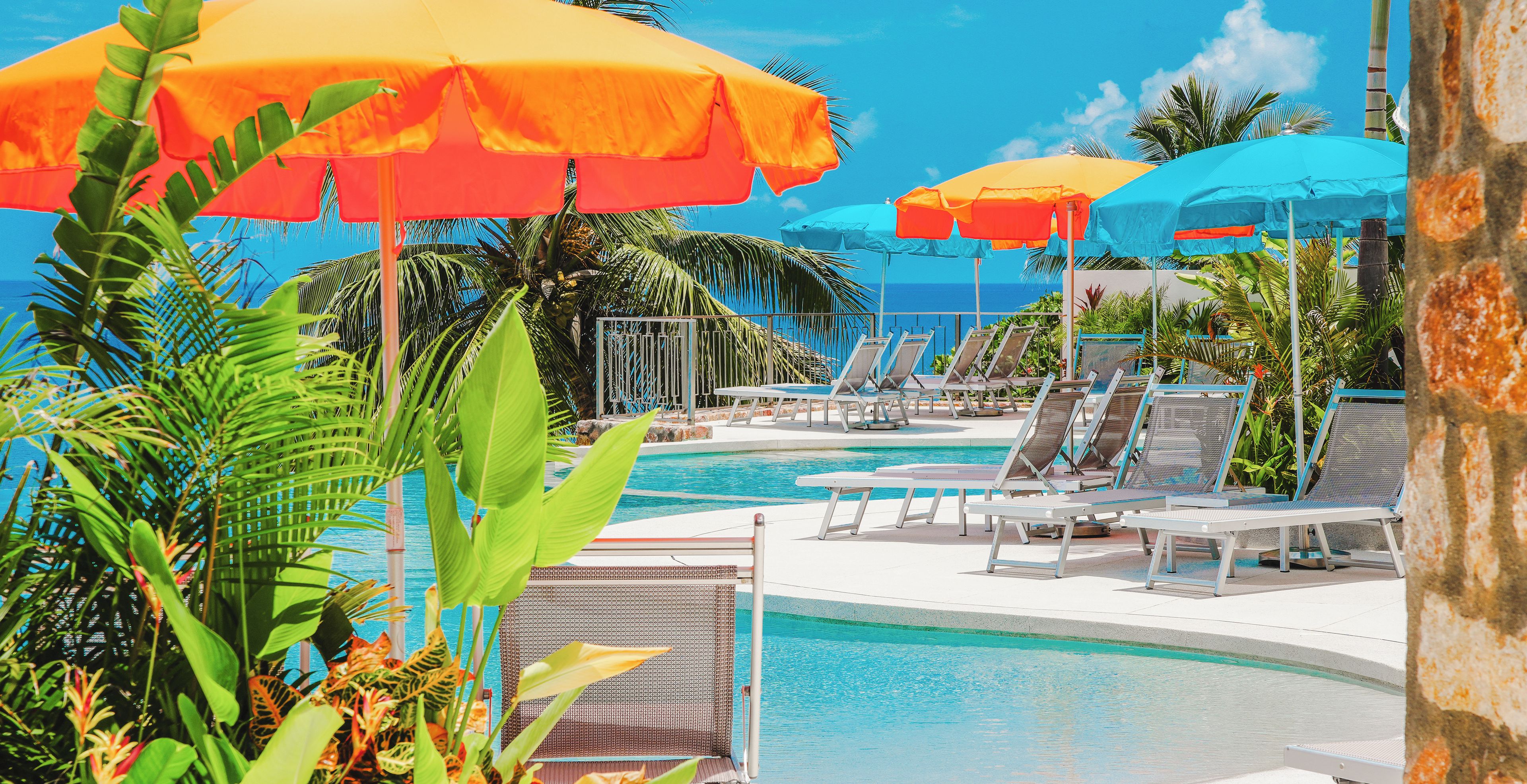 Uma área à beira da piscina com espreguiçadeiras e guarda-sóis, cercada por plantas tropicais.