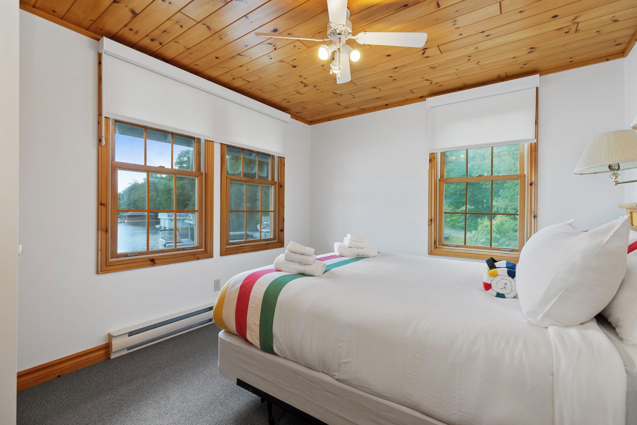 Quarto com teto de madeira, duas janelas com molduras de madeira, uma cama com roupa de cama branca, um aquecedor sob a cama e uma luminária na mesa de cabeceira.