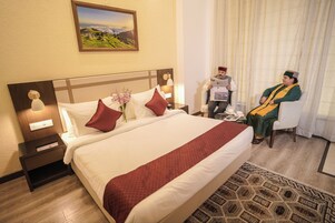 Quarto de hotel com uma cama grande, uma mesa de cabeceira, um abajur, um quadro na parede e uma janela com cortinas.