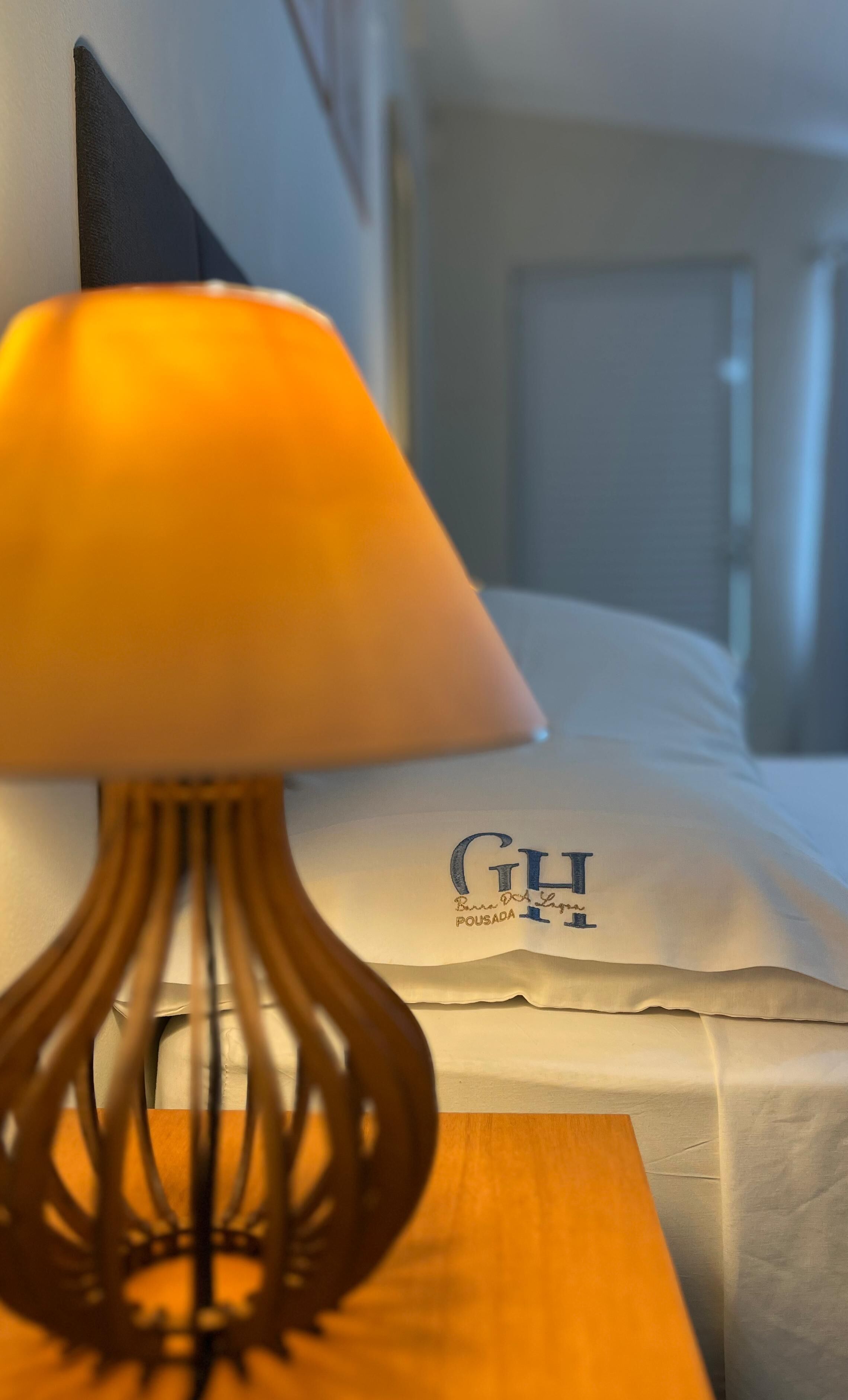 Uma cama com travesseiro exibindo um logotipo e texto, uma mesinha de cabeceira com luminária, e uma janela ao fundo.