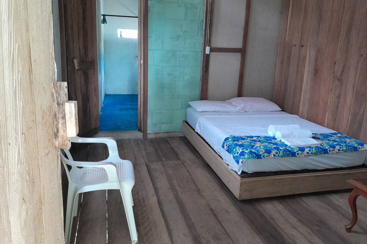 Quarto individual com uma cama, um tapete de madeira, uma cadeira de plástico branca e uma colcha de flores.