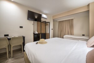 Quarto de hotel com cama, escrivaninha, duas cadeiras, televisão e guarda-roupa.