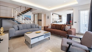 Uma sala de estar moderna com um sofá cinzento, um sofá castanho, uma mesa de centro branca e uma poltrona estampada. Há uma escada com corrimão que leva a um andar superior.