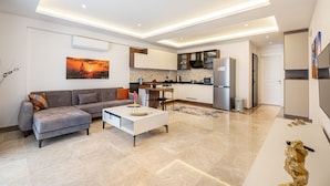 Uma sala de estar moderna com um sofá, mesa de centro e uma pintura de pôr do sol. Ao lado, encontra-se uma cozinha com área de jantar, frigorífico e micro-ondas.
