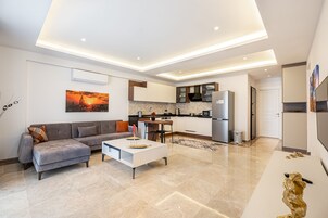 Uma sala de estar moderna com um sofá, mesa de centro e uma pintura de pôr do sol. Ao lado, encontra-se uma cozinha com área de jantar, frigorífico e micro-ondas.