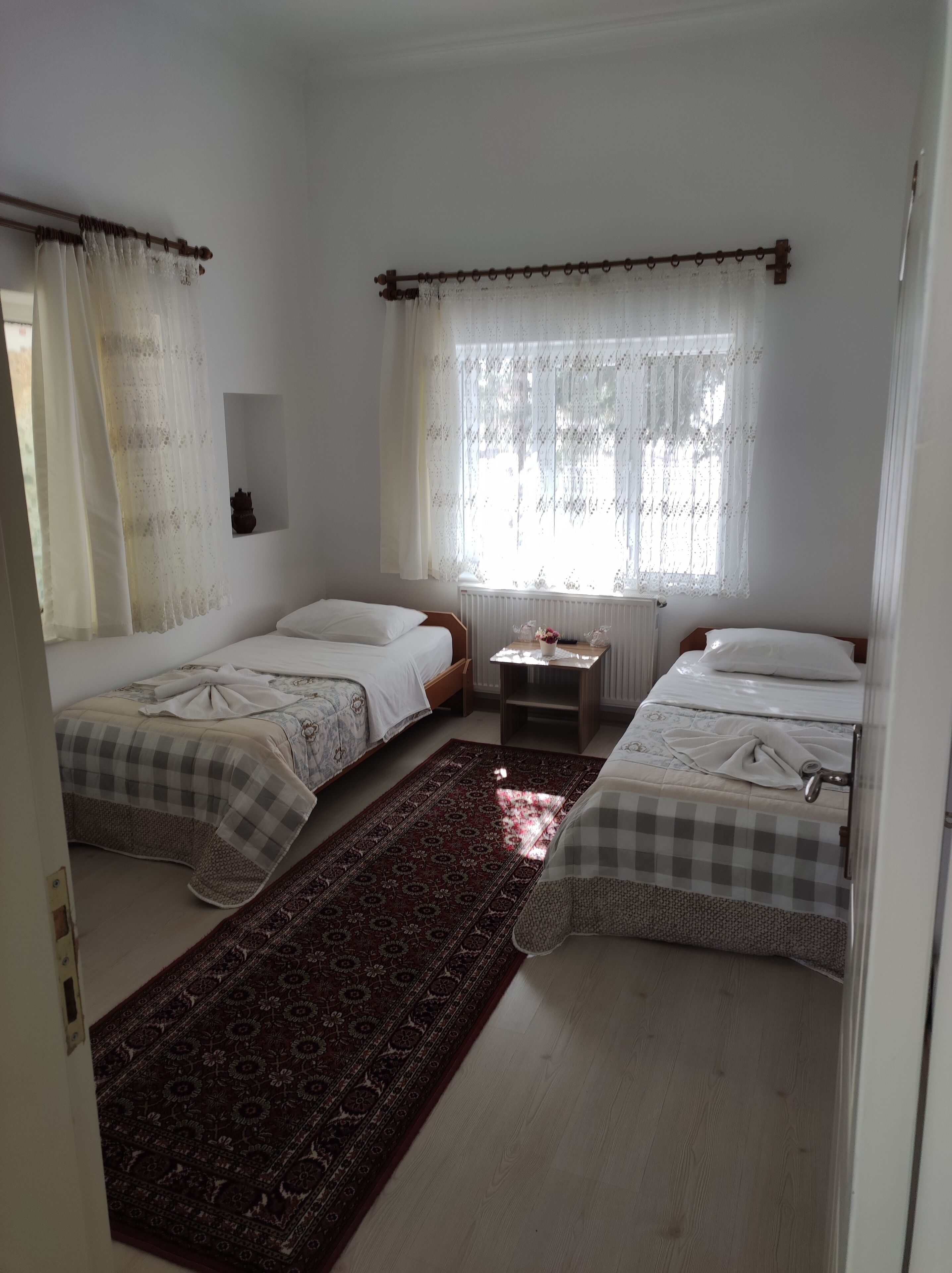 Quarto com duas camas de solteiro, uma mesinha com um vaso, janela com cortinas leves e um tapete escuro com padrão.