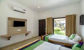 Quarto de hotel com duas camas, uma televisão fixada num painel de madeira, uma porta e vista para um jardim através de uma janela com cortinas.