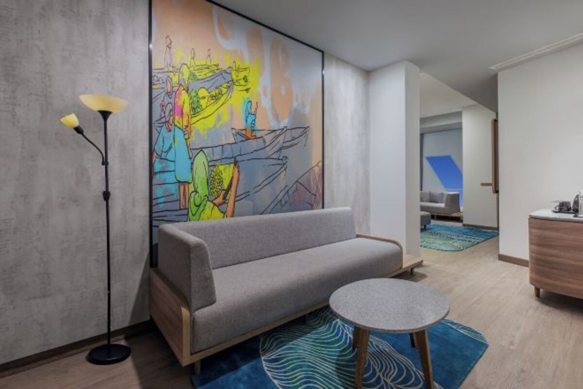 Quarto de hotel moderno com sofá cinzento, mesa redonda, luminária de chão e uma pintura abstrata colorida na parede.